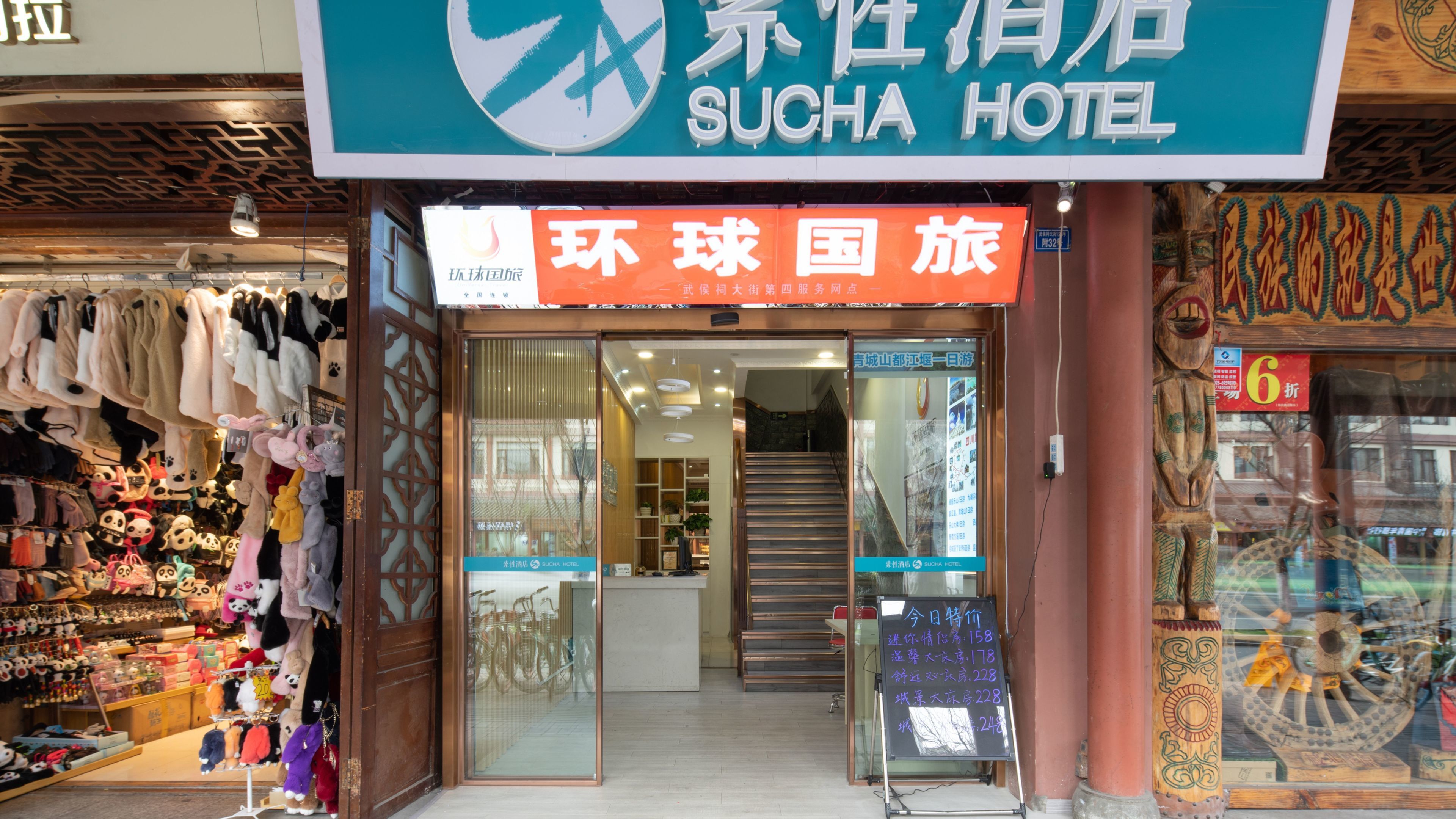 Entrada de hotel com placa da Sucha Hotel, porta de vidro e escadaria que dá acesso ao interior.