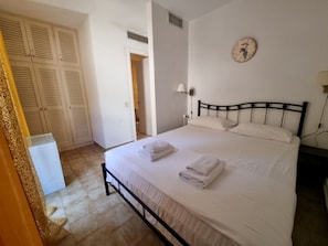 Cama arrumada com toalhas dobradas, relógio de parede e abajur em uma mesa de cabeceira em quarto com piso de azulejos e porta que dá para um armário.