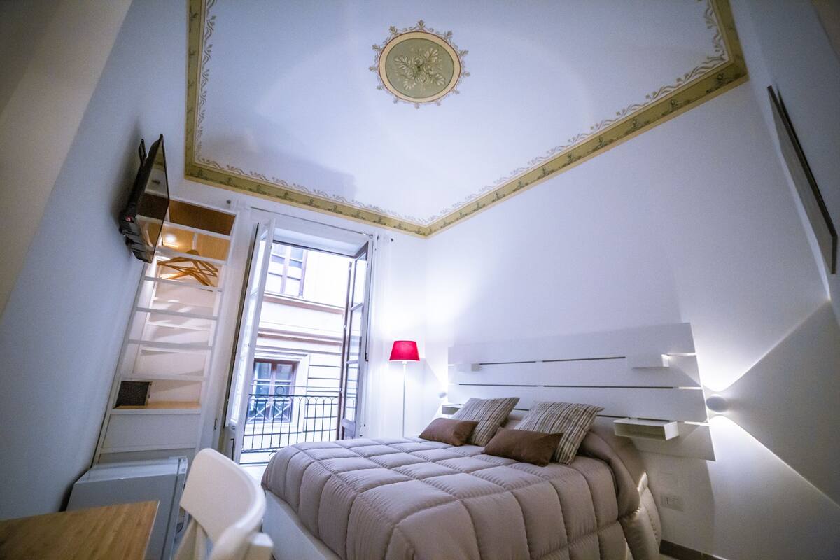 Quarto com cama, escrivaninha, cadeira, janela e uma moldura no teto.