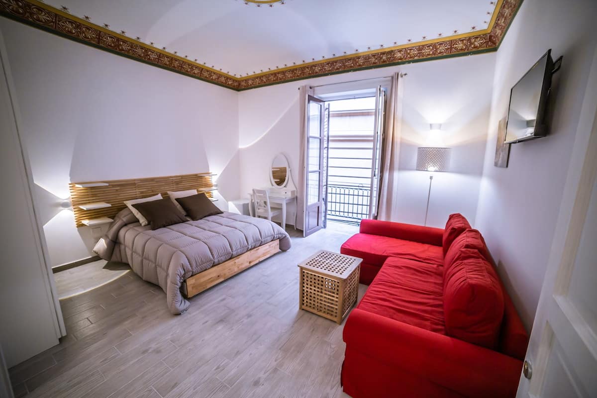 Quarto com cama, sofá vermelho, cabeceira de madeira, mesinha com espelho e janela com vista.