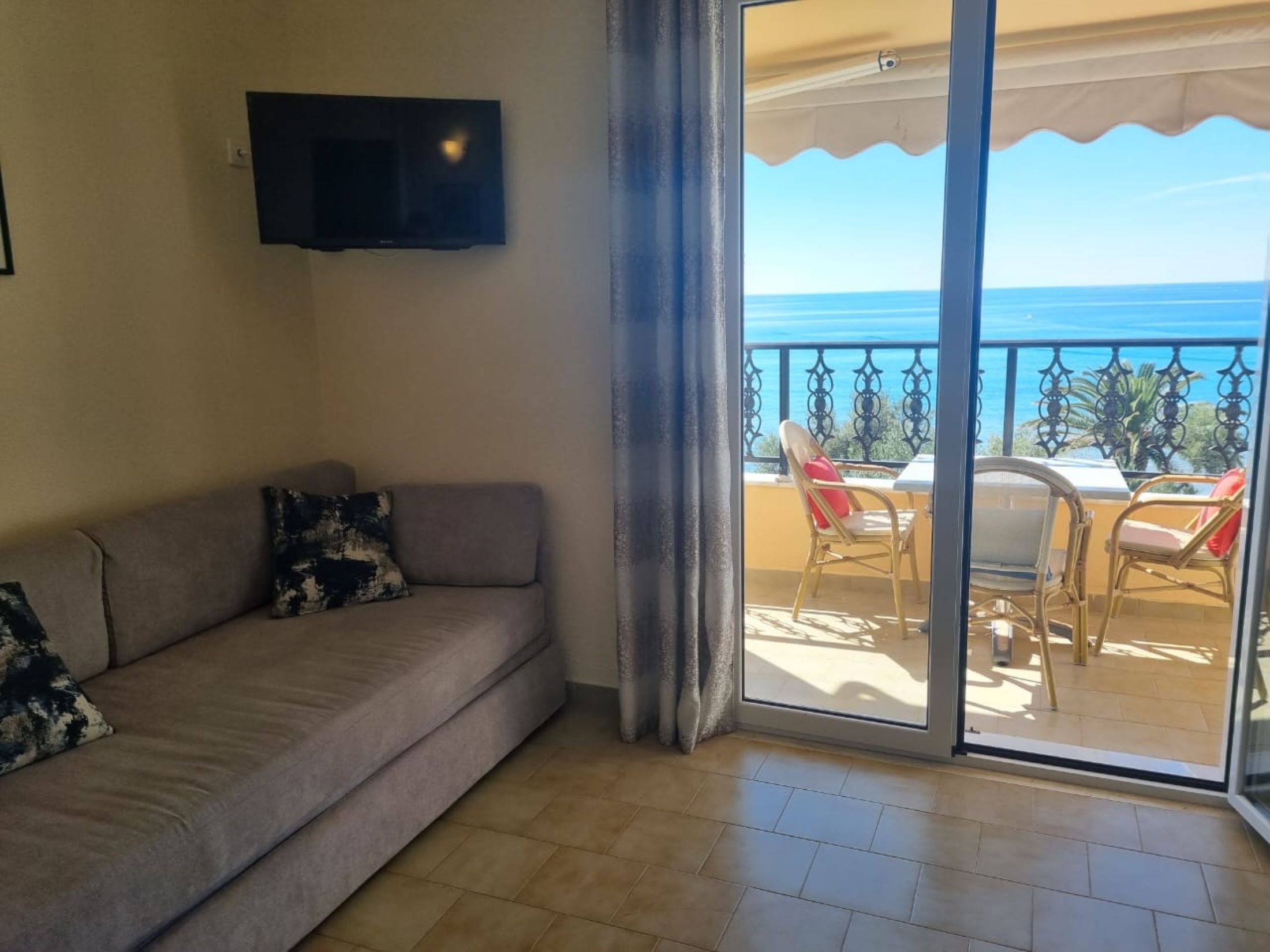 Uma sala de estar com sofá e televisão, uma varanda com cadeiras e mesa, e vista para o mar.