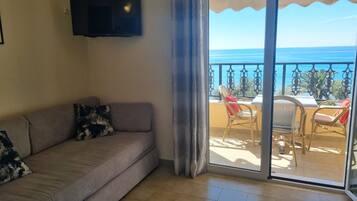 Uma sala de estar com sofá e televisão, uma varanda com cadeiras e mesa, e vista para o mar.