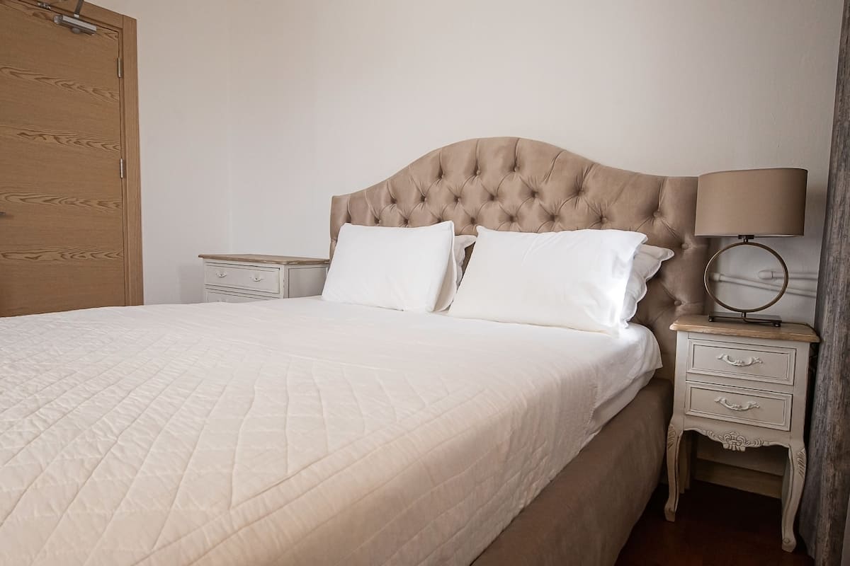 Uma cama bem arrumada com um edredom branco acolchoado, uma cabeceira estofada e duas mesas de cabeceira com luminárias e gavetas.