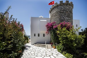 Uma trilha de pedra leva a um edifício branco com uma torre de pedra, cercado por vegetação exuberante e flores cor-de-rosa.