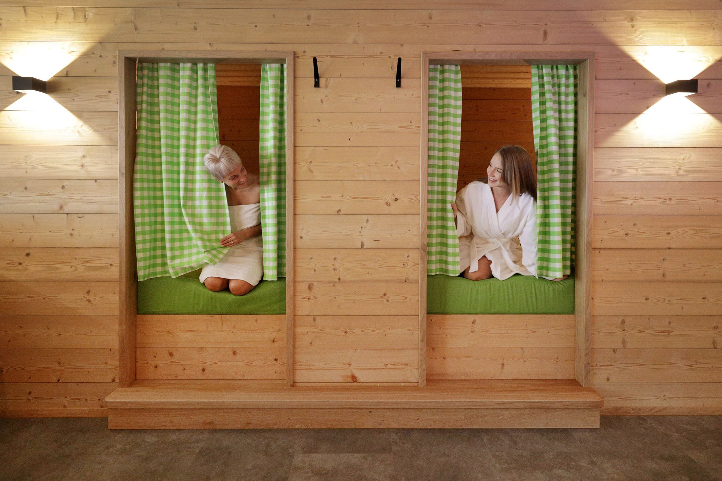 Duas cabanas de madeira com cortinas quadriculas verdes, cada uma com um banco e luzes fixadas na parede.