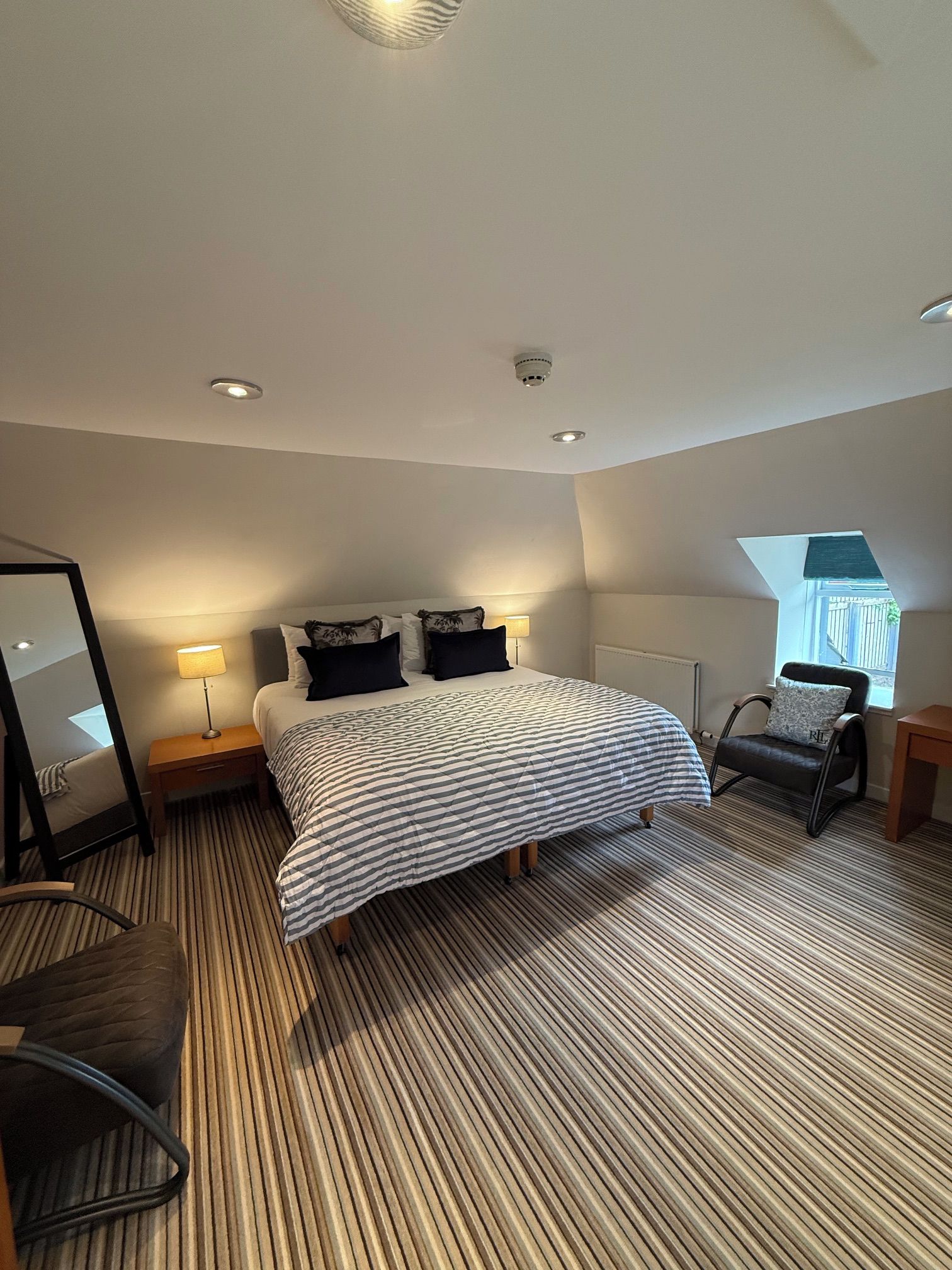 Quarto de hotel com cama, abajures, uma cadeira, espelho e janela.