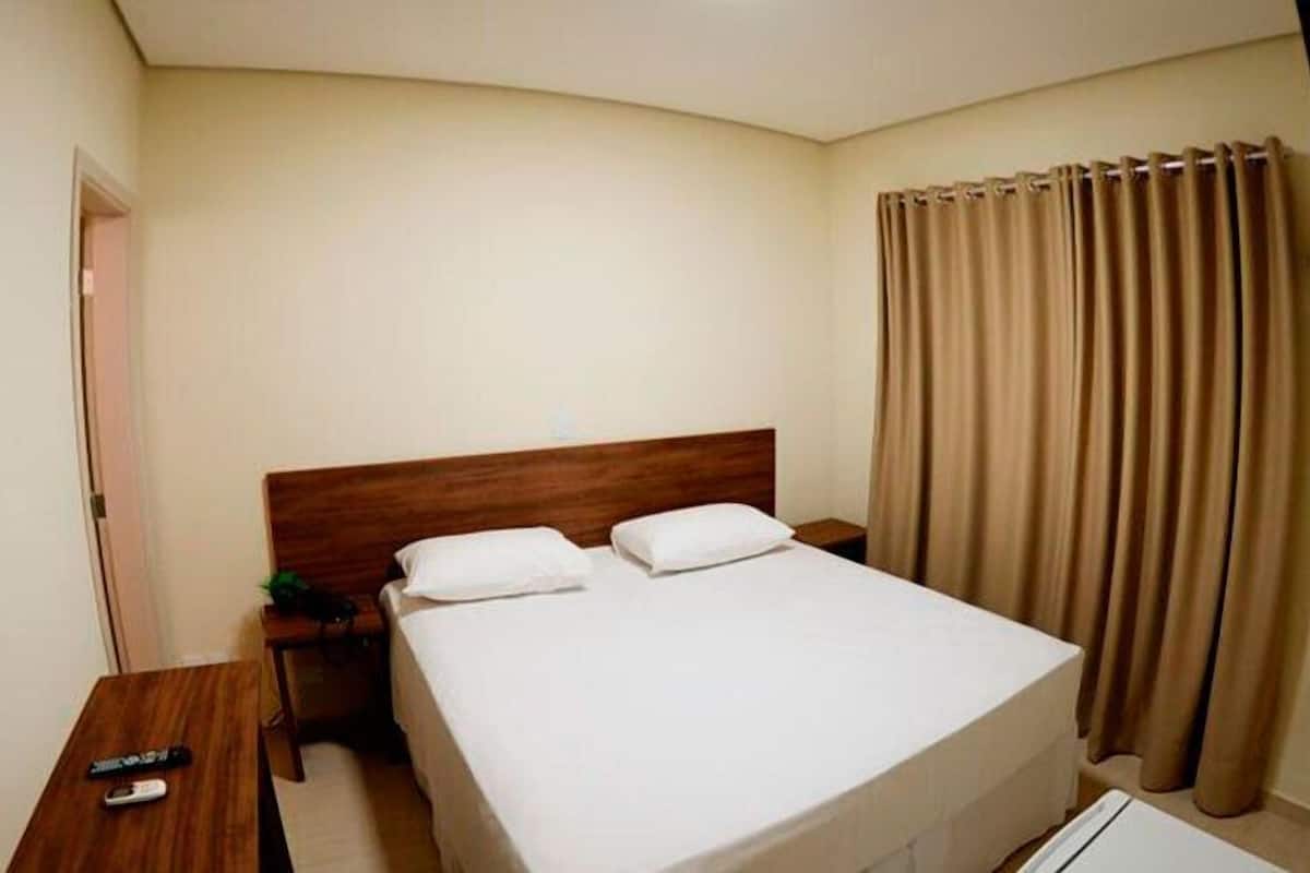 Quarto de hotel com uma cama de solteiro, cabeceira de madeira, mesa de cabeceira com abajur e uma janela com cortinas.