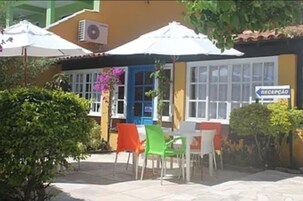 Uma área externa com mesas e cadeiras coloridas e guarda-sóis, uma placa de recepção e um edifício com porta azul e paredes amarelas.