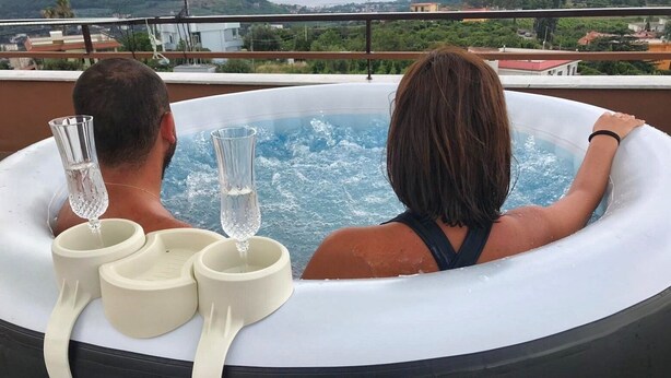 Um casal relaxando em uma banheira de hidromassagem com uma vista panorâmica de colinas e edifícios.