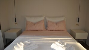 Cama bem arrumada com lençóis brancos e duas almofadas, mesa de cabeceira e luminária fixada na parede.