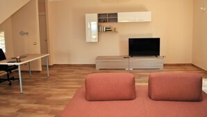 Uma sala de estar moderna com televisão de tela plana, um armário branco com prateleiras, uma escrivaninha com cadeira e uma janela grande.