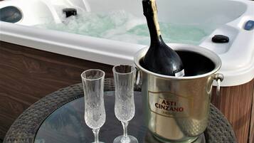 Uma banheira de hidromassagem com uma garrafa de Asti Cinzano e duas taças sobre uma mesa de vidro, com vista para uma paisagem com casas e montanhas.