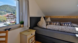 Um quarto aconchegante com uma cama, uma pequena mesa de cabeceira de madeira, uma janela com vista para uma montanha e edifícios, e uma cortina.