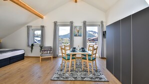 Quarto moderno com teto estilo loft, uma cama, área de jantar com mesa e cadeiras, e vista para montanhas através de grandes janelas.