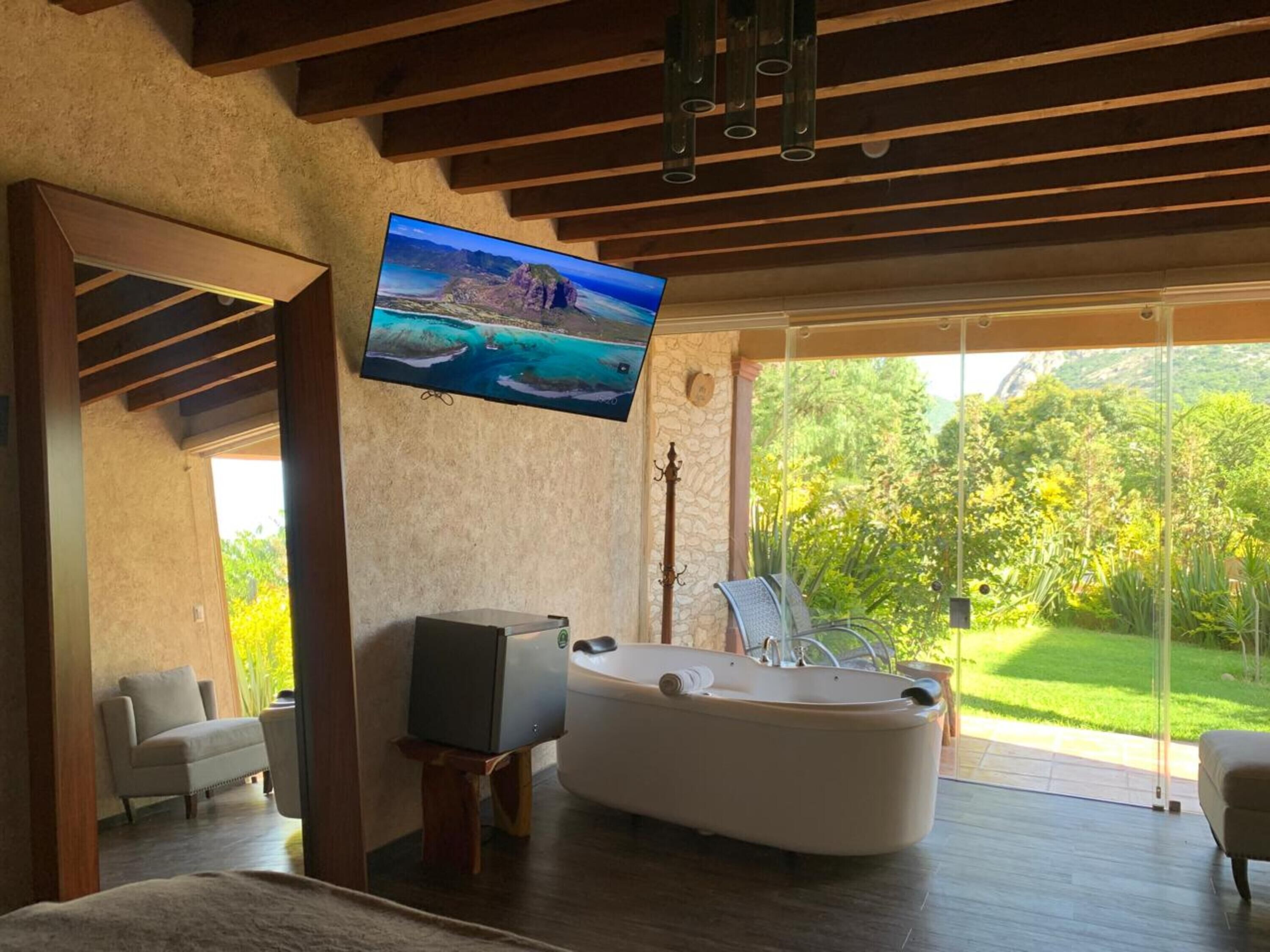 Um quarto espaçoso com uma banheira grande, uma televisão fixada na parede e vista para um jardim exuberante através de amplas portas de vidro deslizantes.