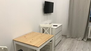 Quarto pequeno e limpo com uma mesa de madeira, uma cadeira branca, uma televisão presa à parede e uma janela com cortina.
