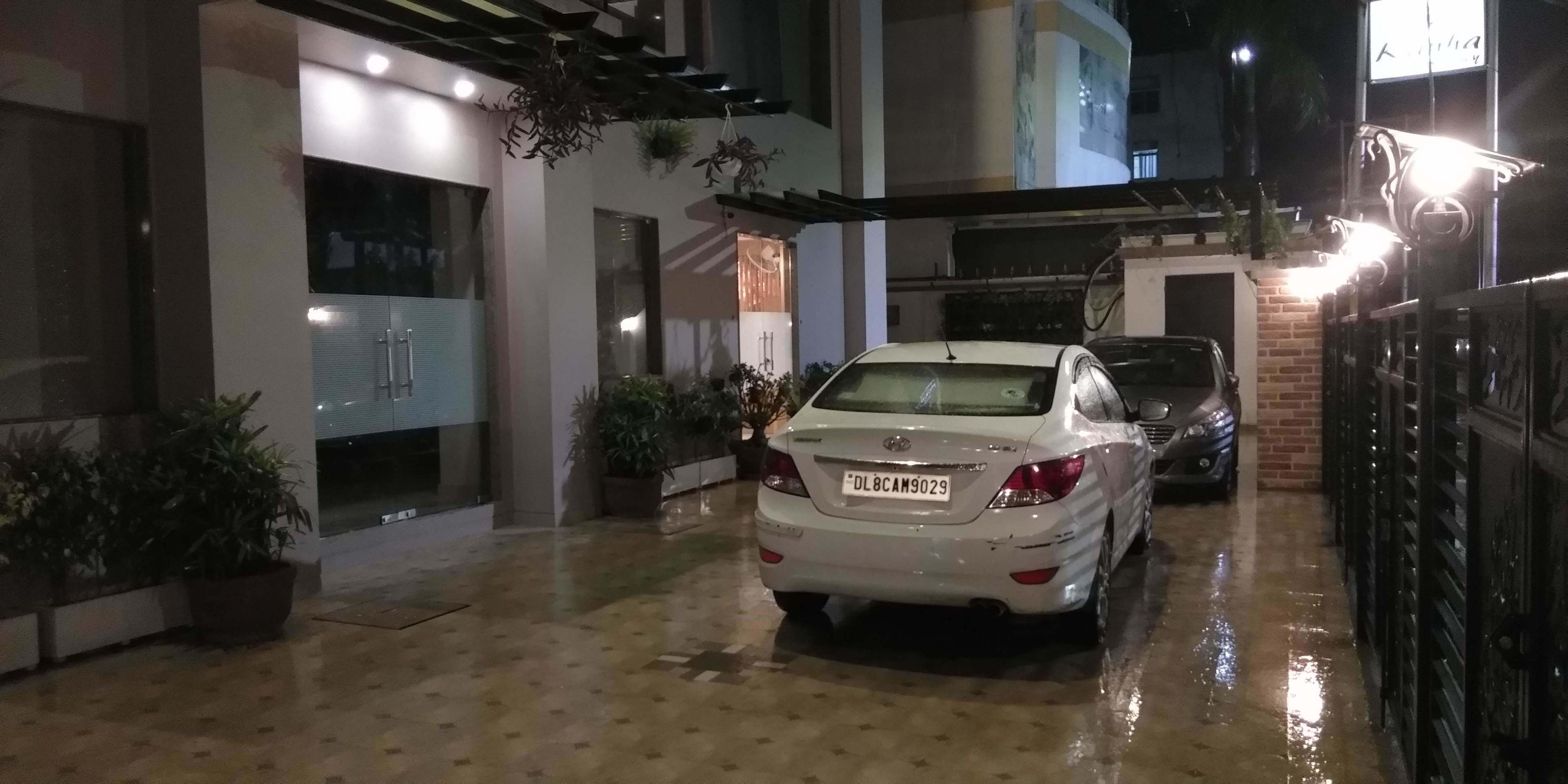 Um carro branco estacionado em área coberta de estacionamento, com plantas em vasos e um edifício moderno ao fundo.