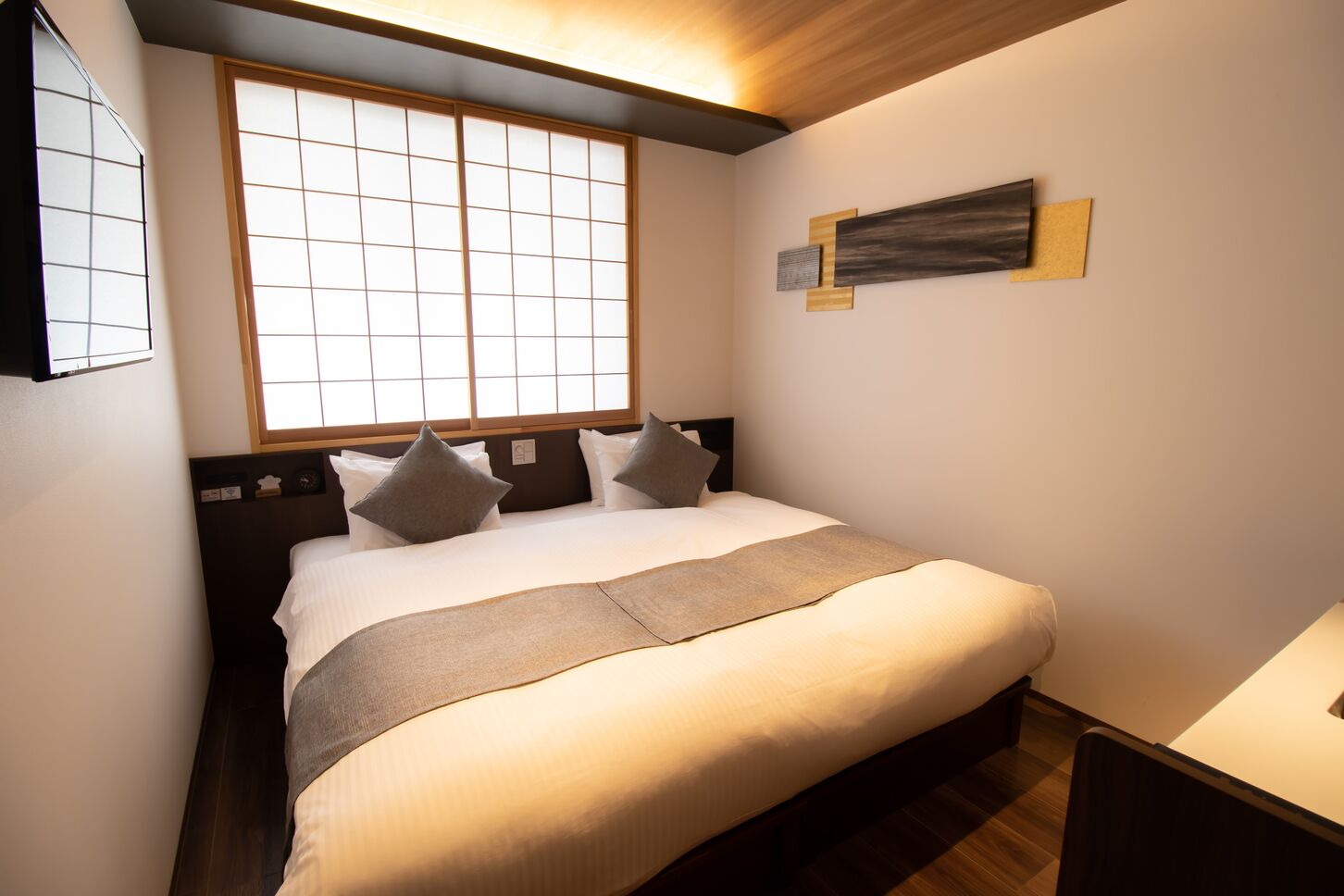 Cama bem arrumada com cabeceira, janela com padrão quadriculado e uma obra de arte na parede.