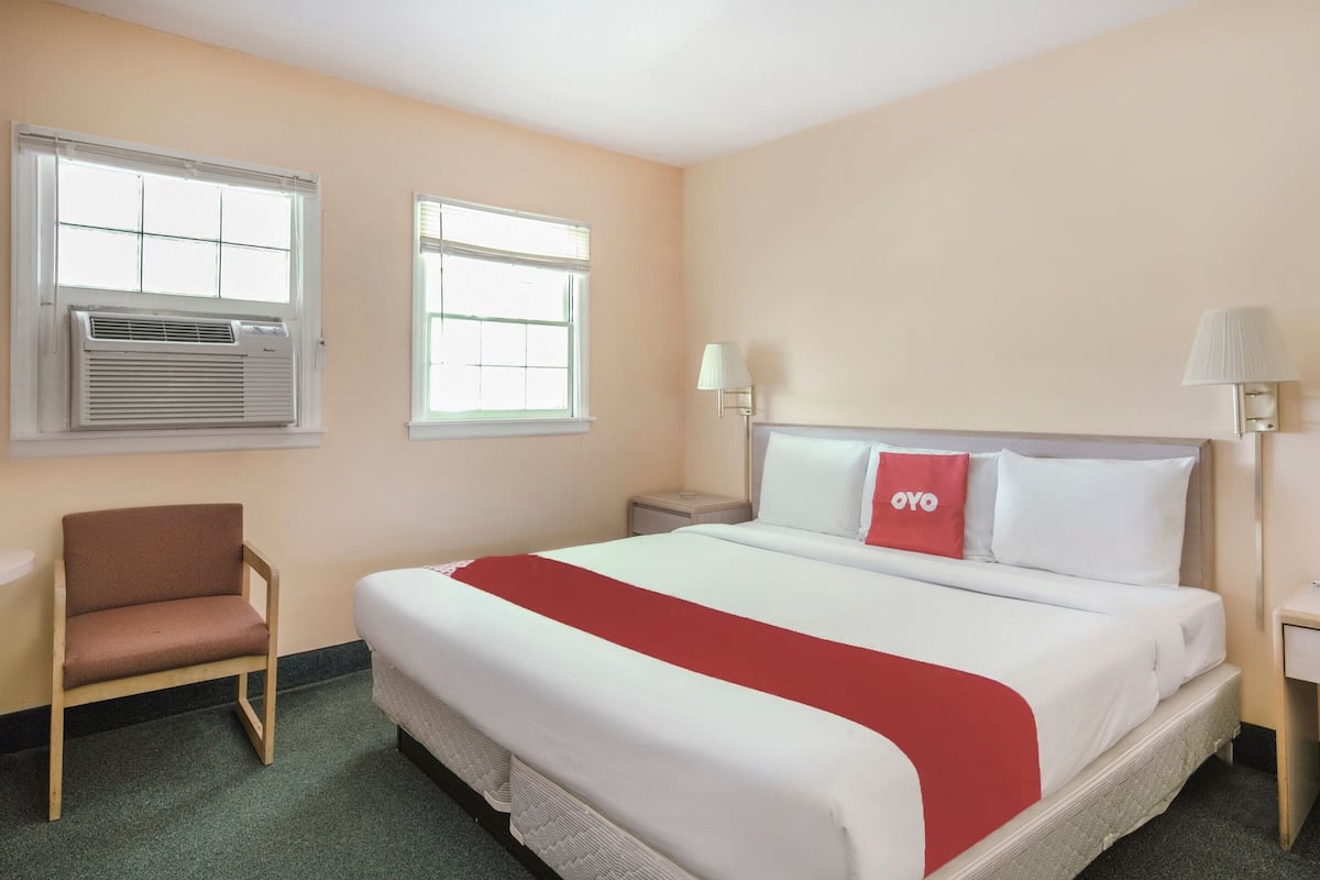 Quarto de hotel com uma cama grande, duas mesas de cabeceira, uma cadeira e duas janelas com persianas.