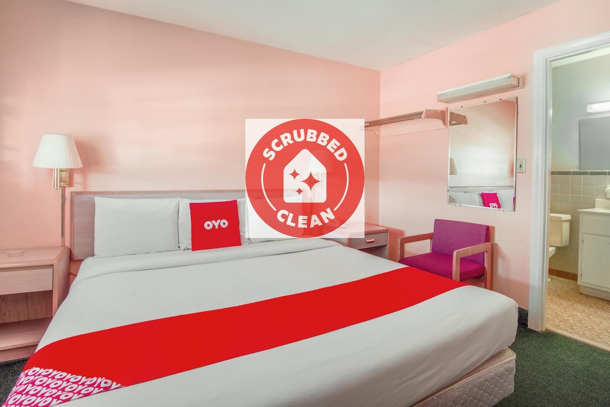 Quarto de hotel com cama, mesa de cabeceira, luminária, cadeira, espelho e banheiro visível através de uma porta aberta.