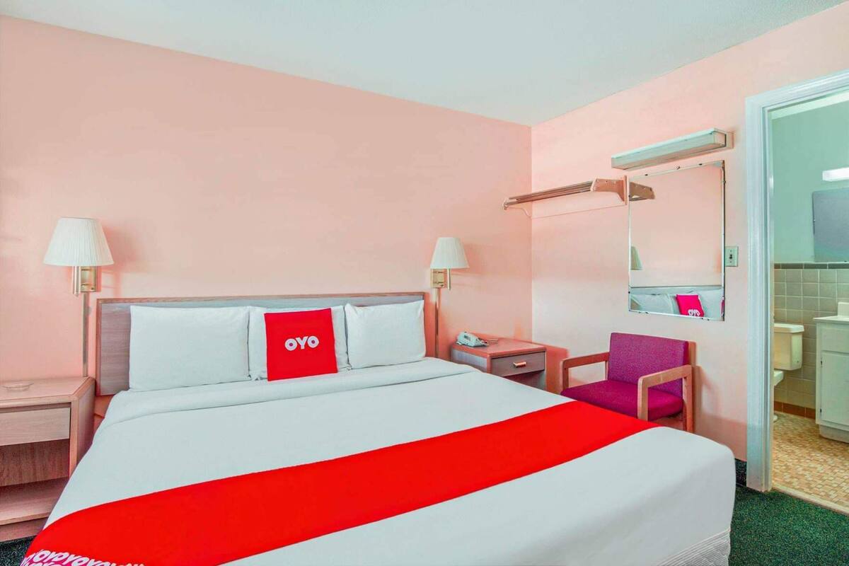 Quarto de hotel com cama, mesas de cabeceira, uma cadeira, um espelho e banheiro visível por uma porta aberta.