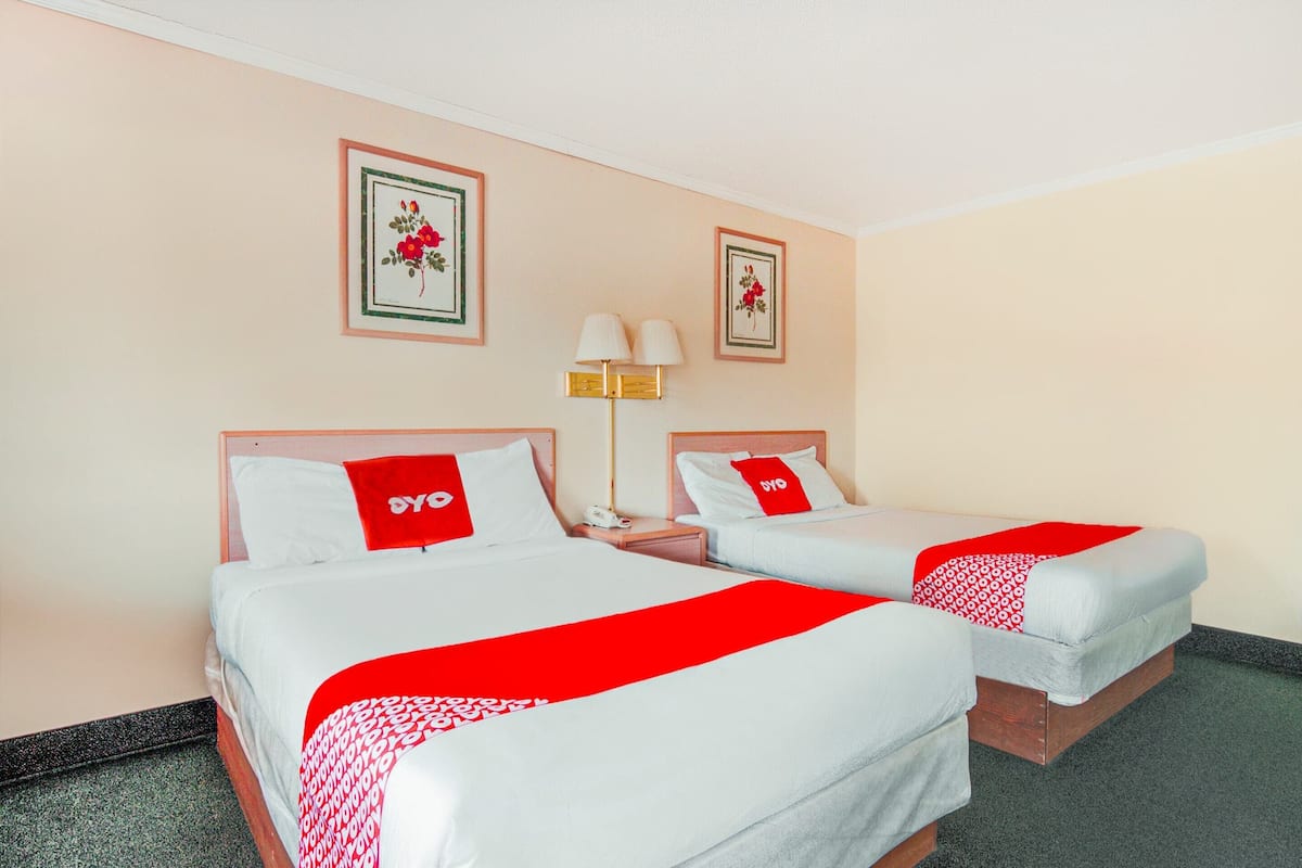 Quarto de hotel com duas camas, cada uma com colcha vermelha e branca e almofadas combinando. Há quadros com flores nas paredes, uma mesa de cabeceira com abajur e um telefone.
