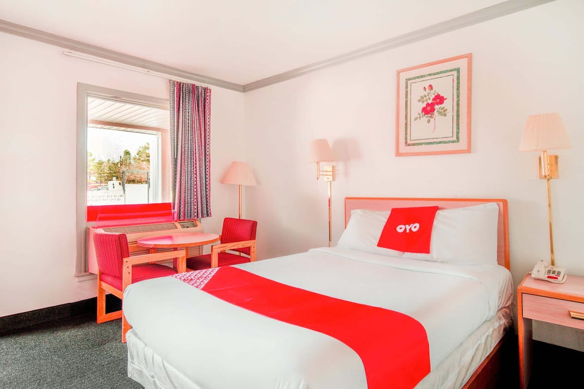 Quarto de hotel com cama, escrivaninha, cadeira, janela com cortinas e luminária na parede.