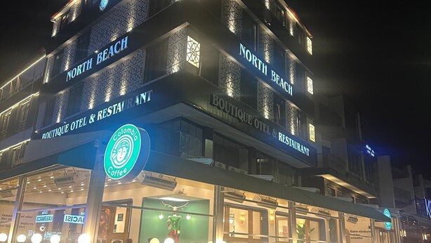 Um edifício de vários andares com a placa 'North Beach Boutique Hotel & Restaurant', exibindo uma placa chamativa 'Colombia Coffee' e uma área de entrada bem iluminada.