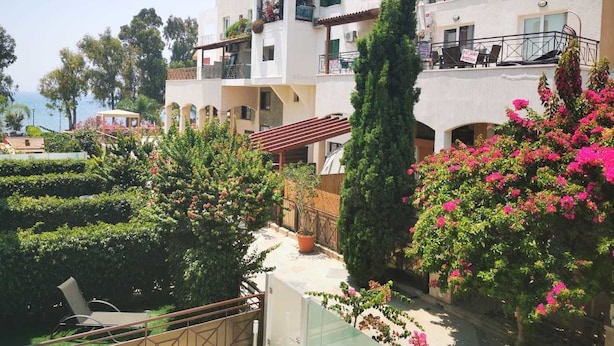 Um edifício de vários andares com varandas, um jardim com sebes e plantas floridas, e vista clara para o mar.