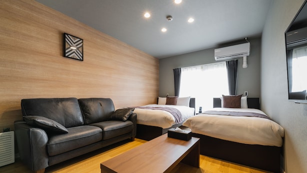 Quarto de hotel moderno com parede de destaque em madeira, sofá de couro preto, mesa de centro de madeira, cama com colcha branca e marrom, e um aparelho de ar condicionado.