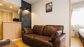 Uma sala de estar moderna com um sofá de couro marrom, piso de madeira, uma pequena cozinha e um quadro na parede.