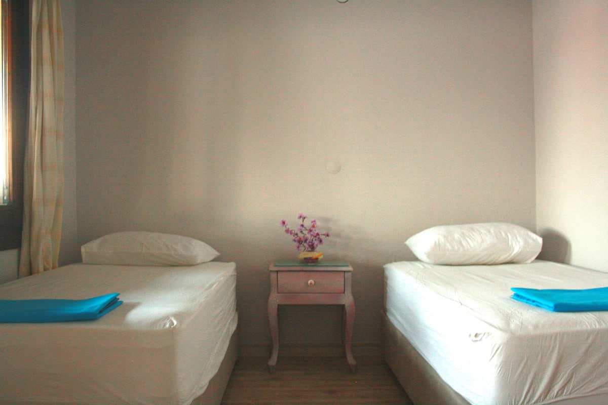 Quarto de hotel com duas camas, uma mesinha de cabeceira de madeira com um vaso de flores e uma janela com cortinas.