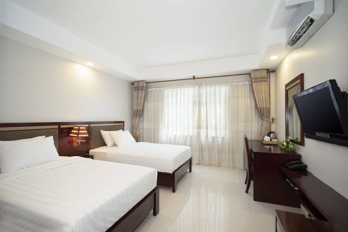 Quarto de hotel com duas camas, uma escrivaninha, televisão e janela com cortinas.