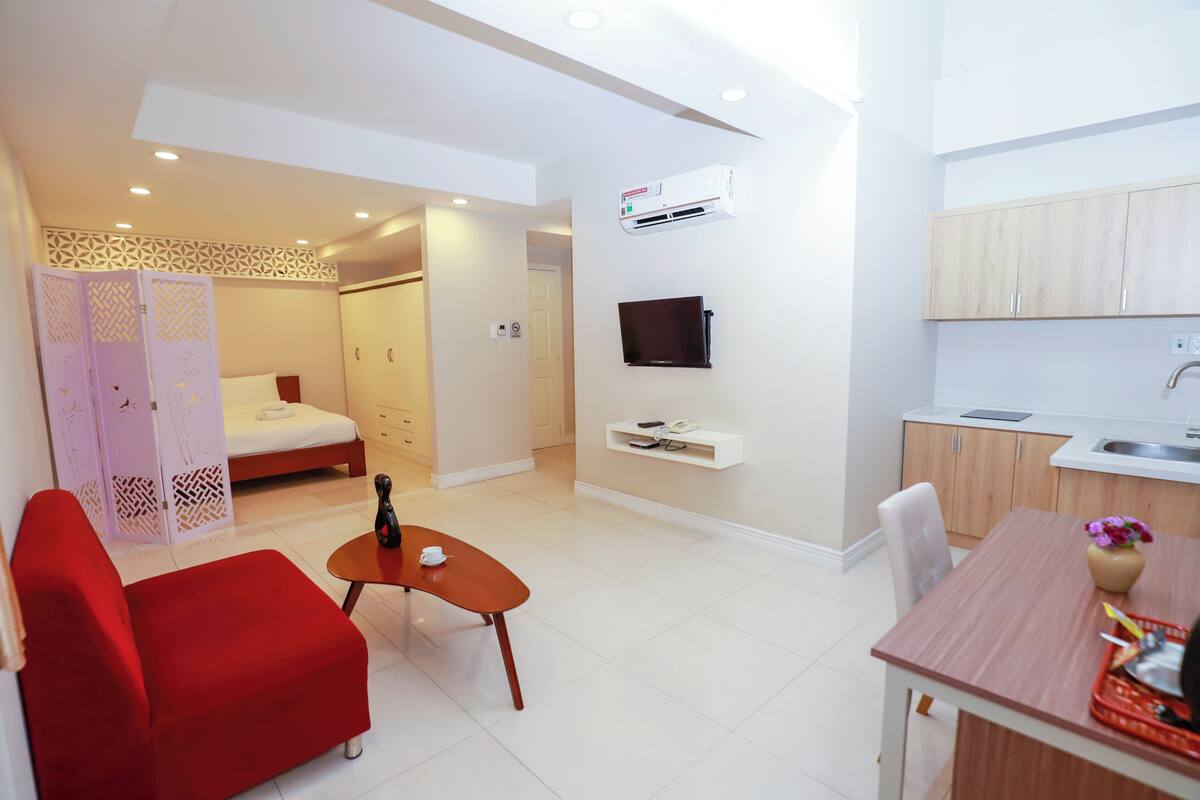 Quarto de hotel moderno com cama, sofá vermelho, pequena mesa de jantar, televisão e cozinha compacta.