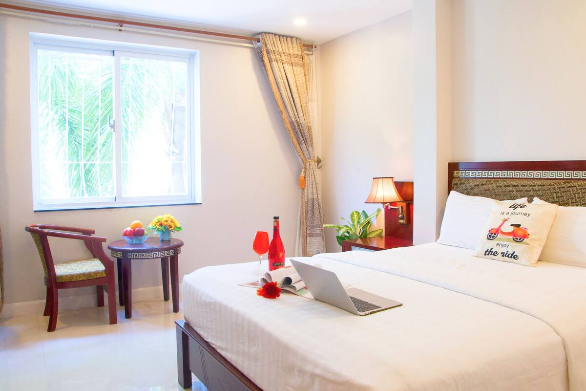 Quarto de hotel com uma cama, uma escrivaninha com um laptop, uma cadeira, uma mesinha com um vaso de flores e uma janela com cortinas.