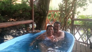Um casal em uma banheira de hidromassagem com deck de madeira e vegetação ao fundo.