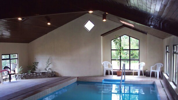 Piscina interna com teto de madeira, janelas amplas e cadeiras brancas ao redor da piscina.