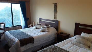 Cama bem arrumada com roupa de cama branca e duas almofadas, mesa de cabeceira com abajur e janela com cortinas.
