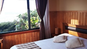 Uma cama bem arrumada com manta listrada e duas almofadas, cabeceira de madeira, luminária pendente e uma janela com cortinas.