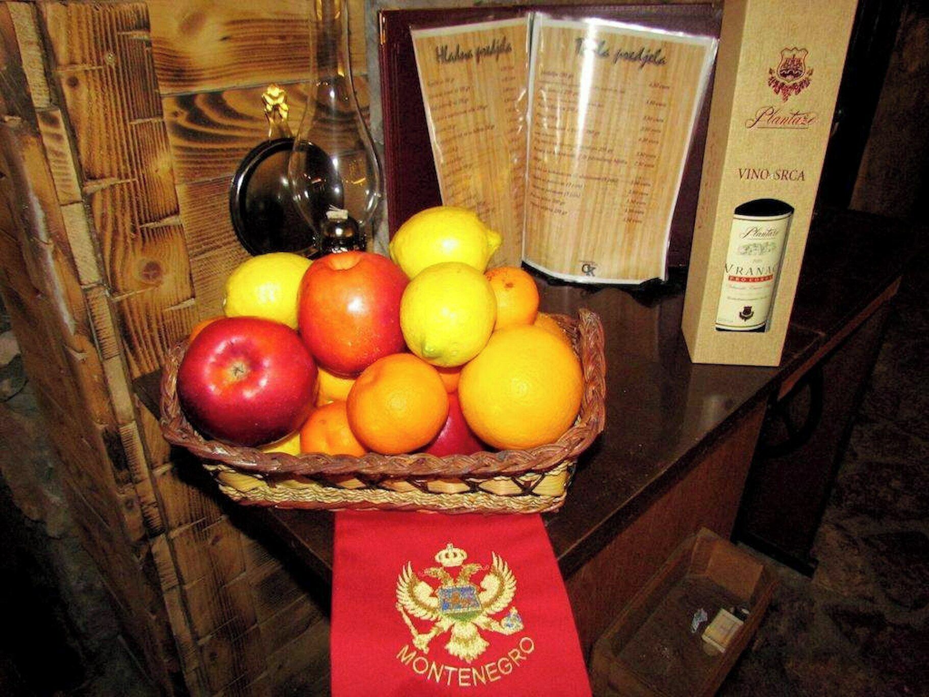 Uma mesa de madeira com uma cesta de frutas, uma garrafa e um menu.