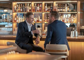 Dois homens de terno sentados em um balcão de bar, com uma prateleira repleta de bebidas ao fundo.