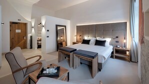 Quarto de hotel moderno com uma cama grande, uma cadeira de madeira, uma mesinha com um tabuleiro e uma mesa de cabeceira com um abajur.