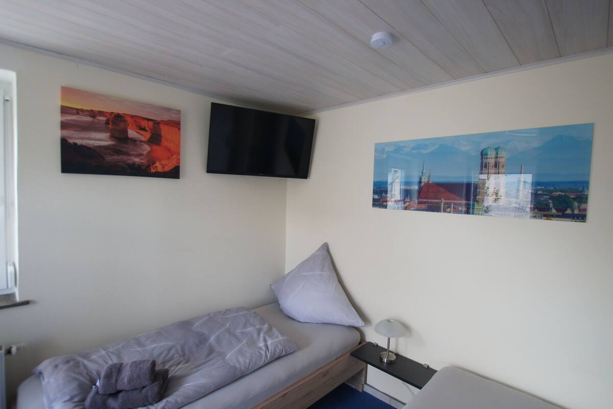 Uma cama de solteiro com estrutura de madeira, um criado-mudo com abajur e duas pinturas penduradas na parede retratando paisagens.
