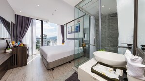 Quarto de hotel moderno com uma cama grande, chuveiro em vidro e pia branca, e vista para a cidade.