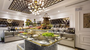 Uma área de pequeno-almoço de hotel moderna, com uma ilha de mármore, diversas opções de comida e um teto decorativo.