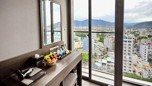 Quarto de hotel com uma janela ampla que oferece vista da cidade, uma escrivaninha de madeira com espelho e uma cesta de frutas.