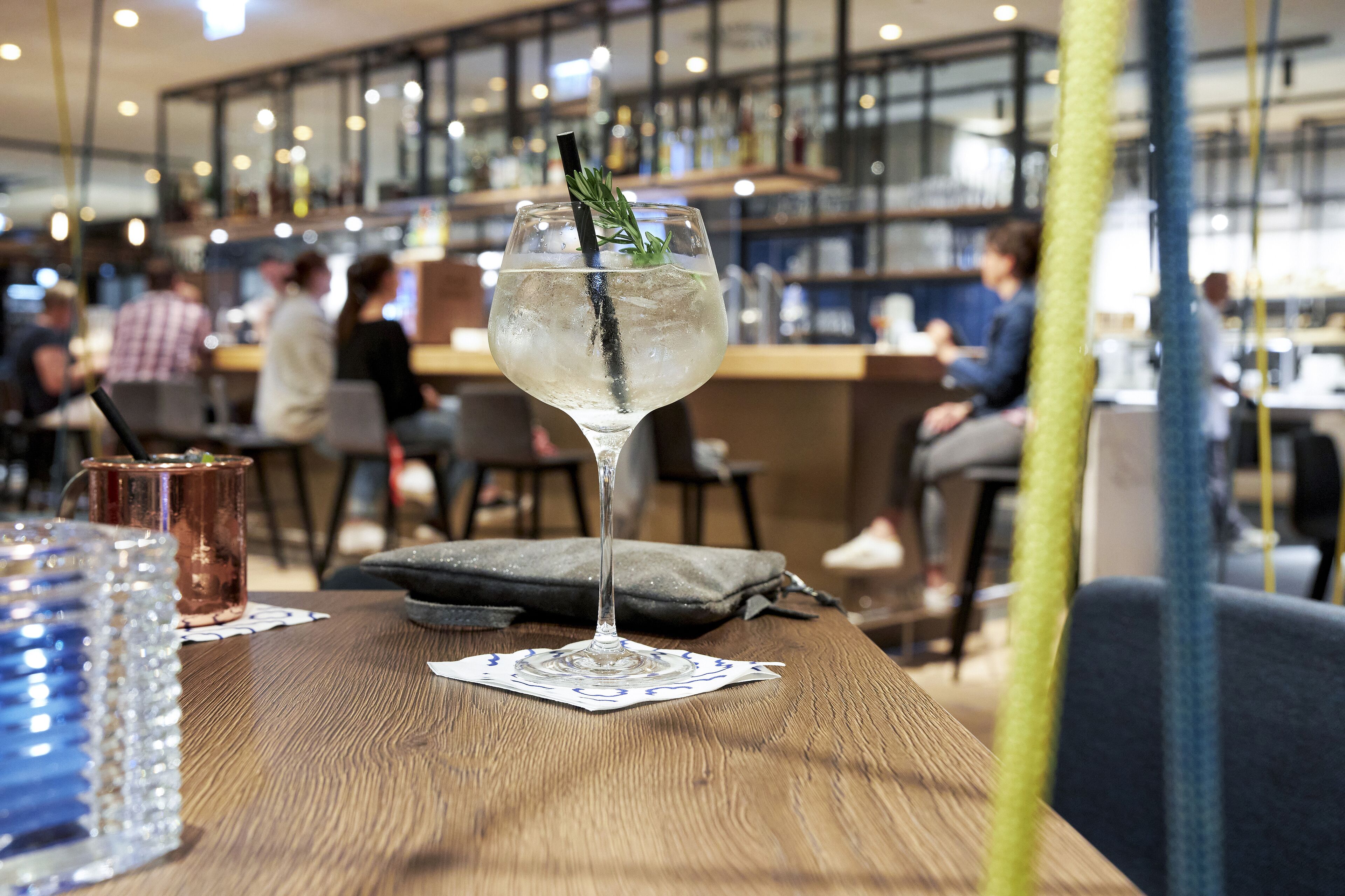 Um copo de vinho branco com um ramo de alecrim sobre uma mesa de madeira num restaurante com decoração moderna e clientes ao fundo.