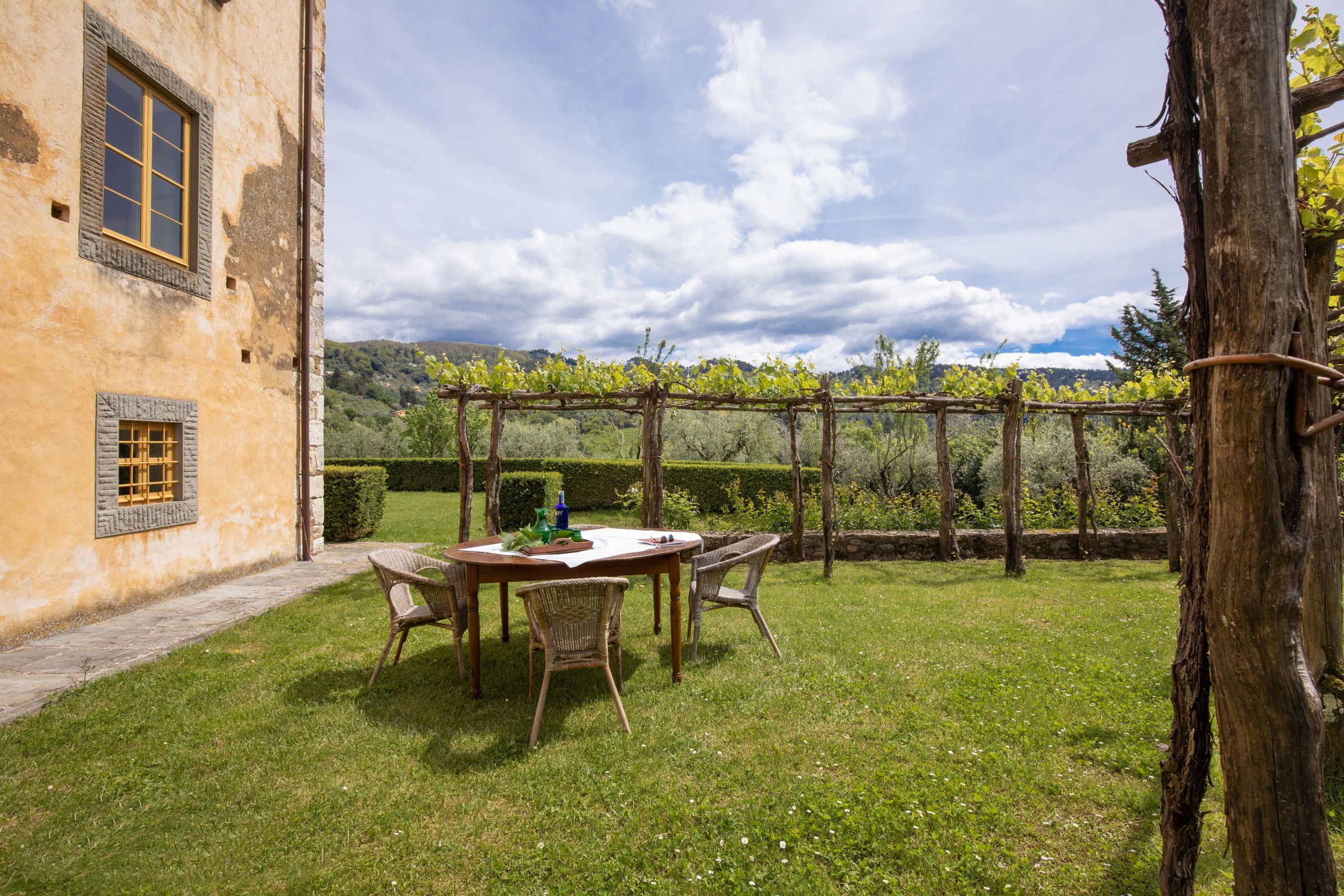 Uma área de refeições rústica ao ar livre, com mesa e cadeiras, cercada por uma pérgola de madeira e vista para vinhedos.