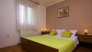 Quarto com cama de madeira, almofadas verdes, colcha branca, mesa de cabeceira com abajur, pintura na parede e janela com cortinas leves.