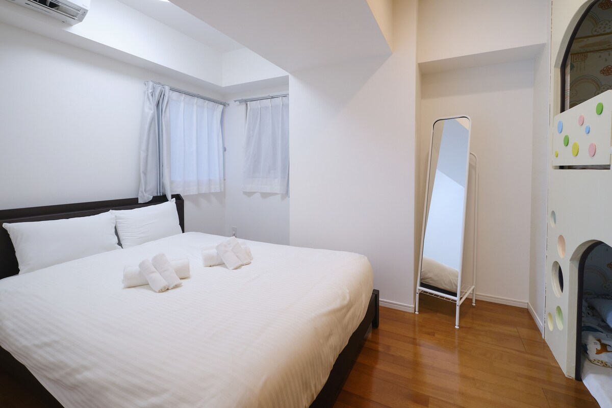 Cama arrumada com lençóis e toalhas brancos, um espelho de corpo inteiro e uma cama beliche infantil com decorações coloridas.
